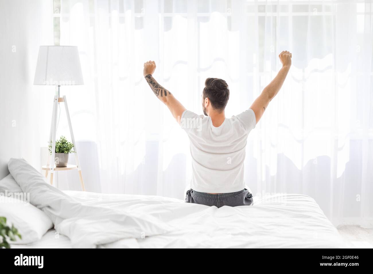 Relaxed young man waking up in bed and stretching his arms, facing the ...