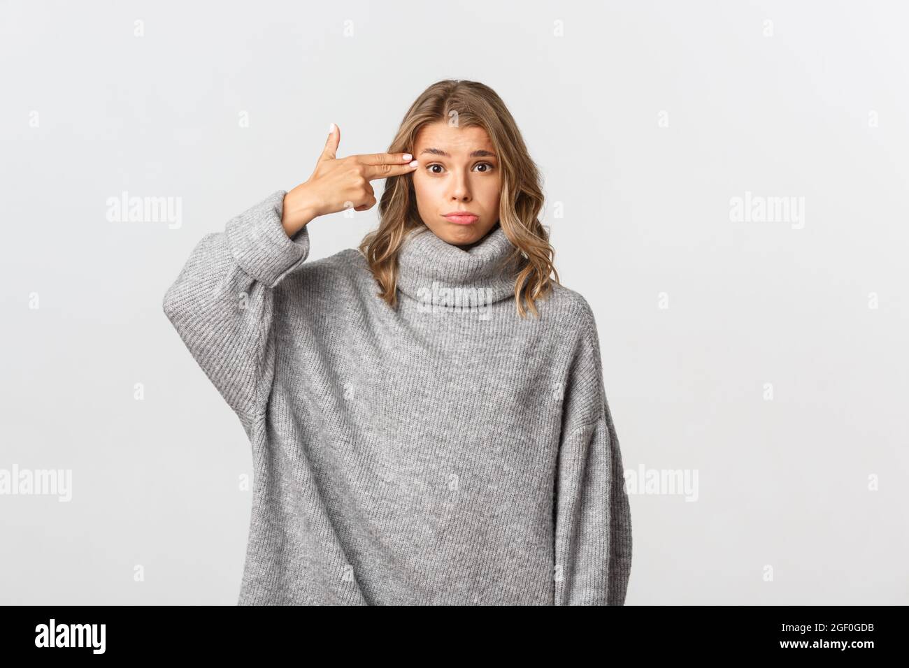 Portrait of sad blond girl in grey sweater, making finger gun sign near ...