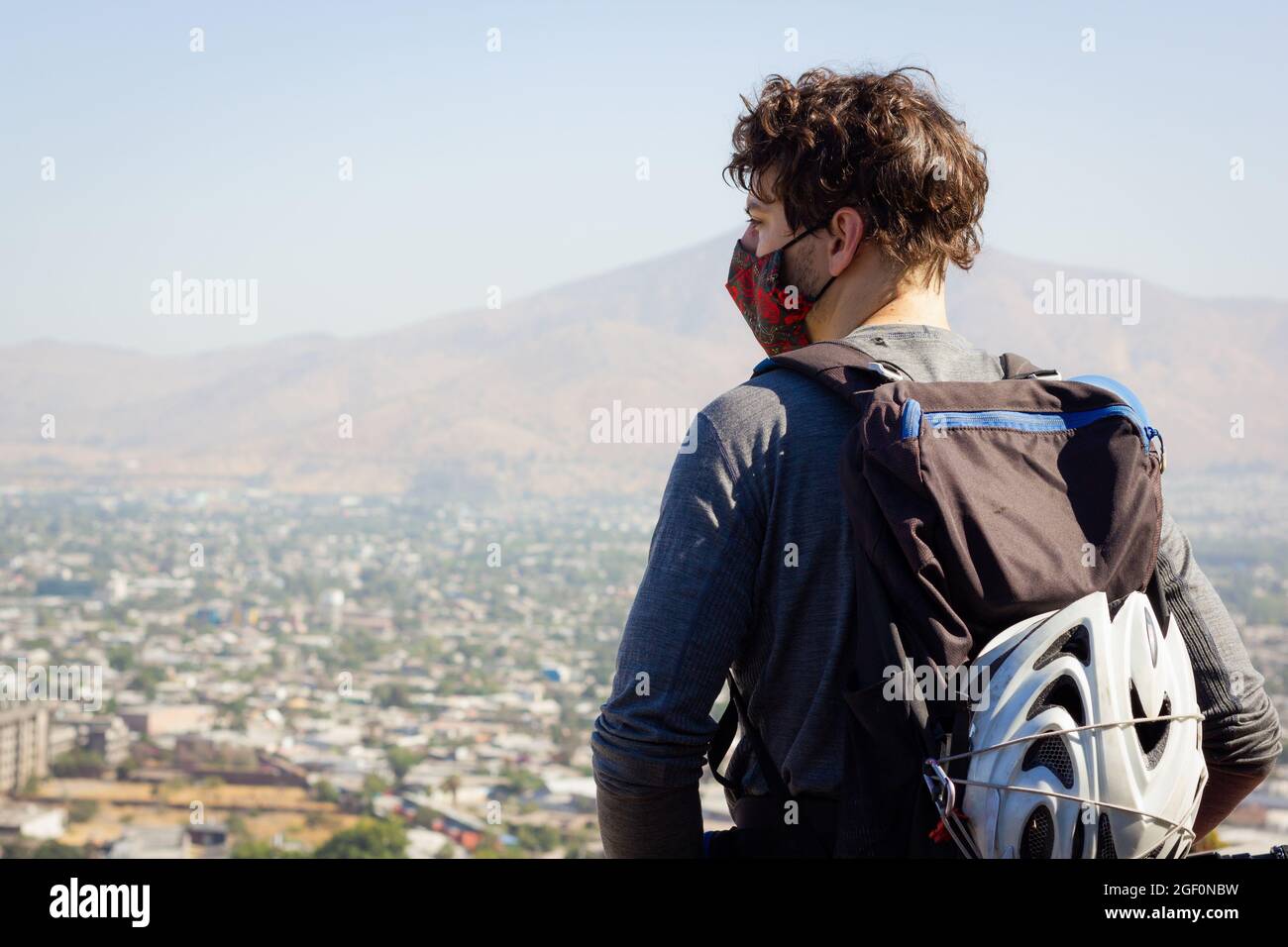 Young biker with face covering and helmet on backpack looking at city ...