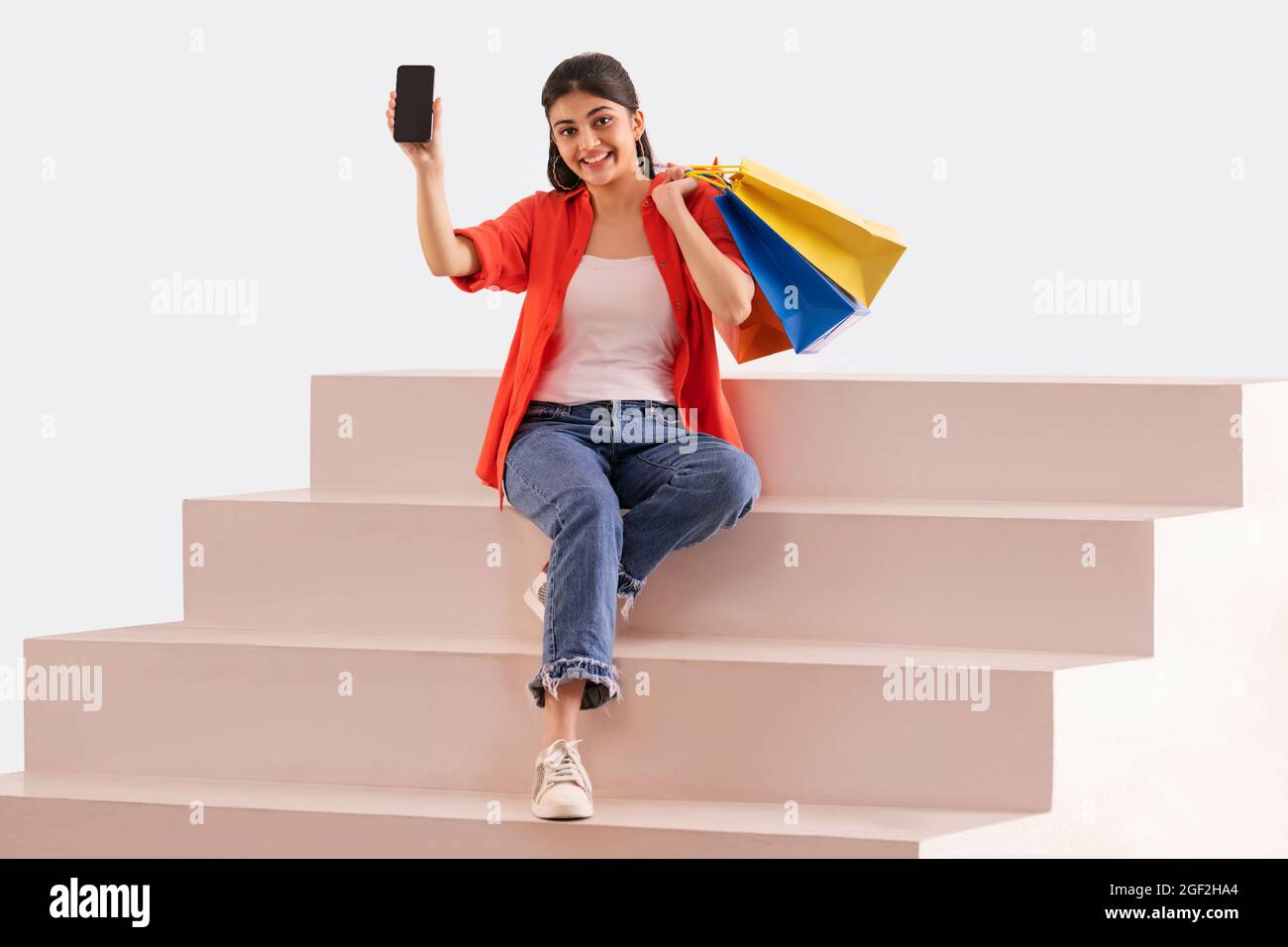 A young woman sitting on stairs holding colorful carrybags showing her ...