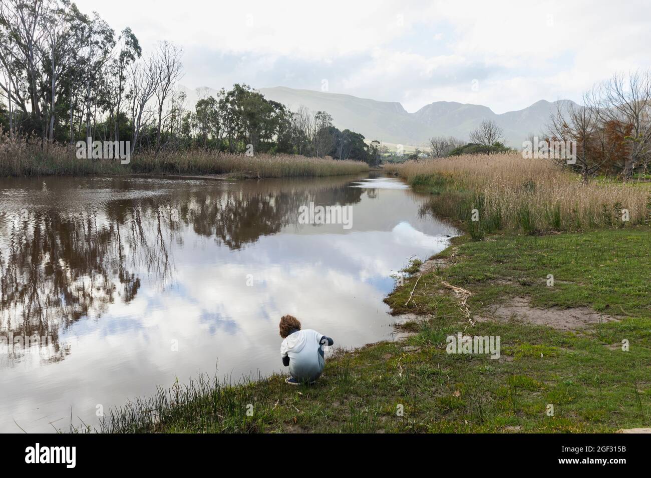 Young boy playing on a river bank, flat calm water and open spaces ...