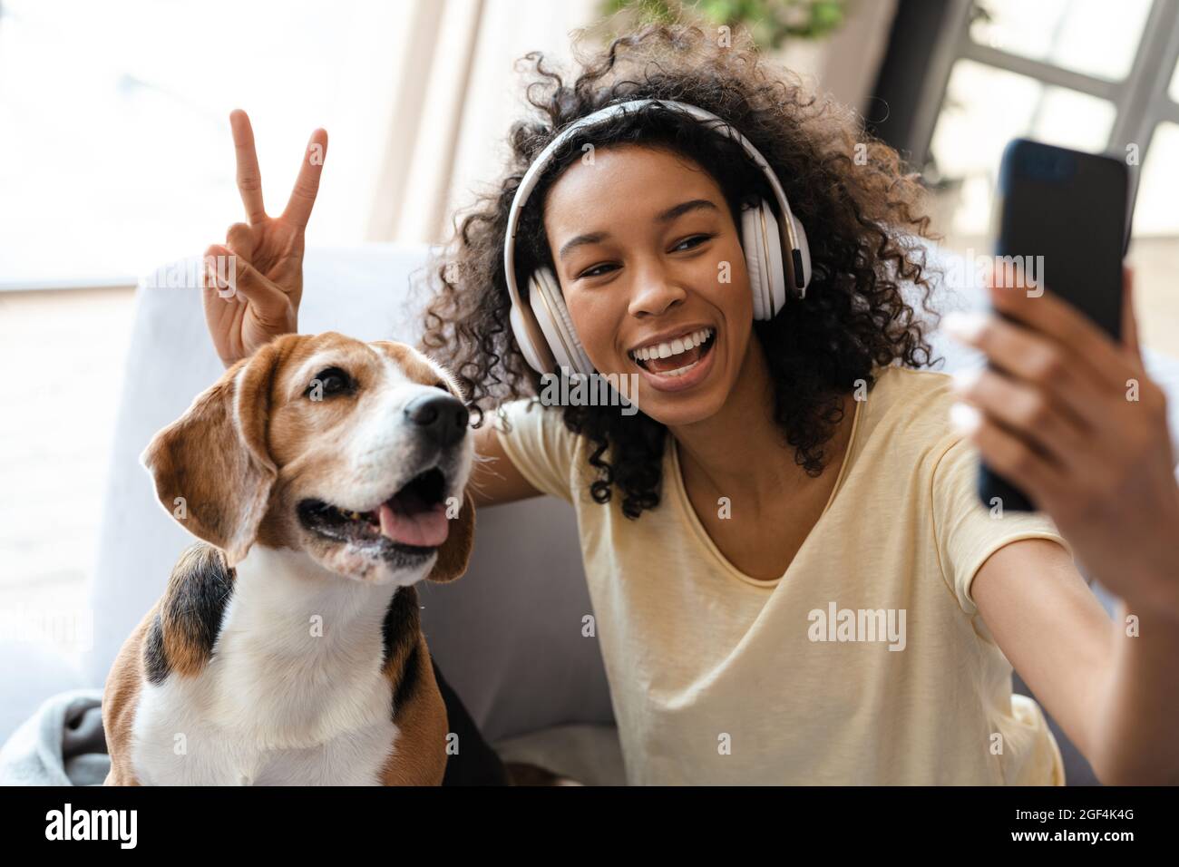 Happy young african woman in headphones relaxing on a couch with her pet dog at home, taing a ...