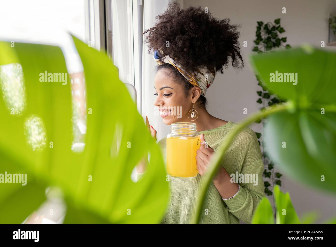 Smiling young woman looking through window while holding juice jar at ...
