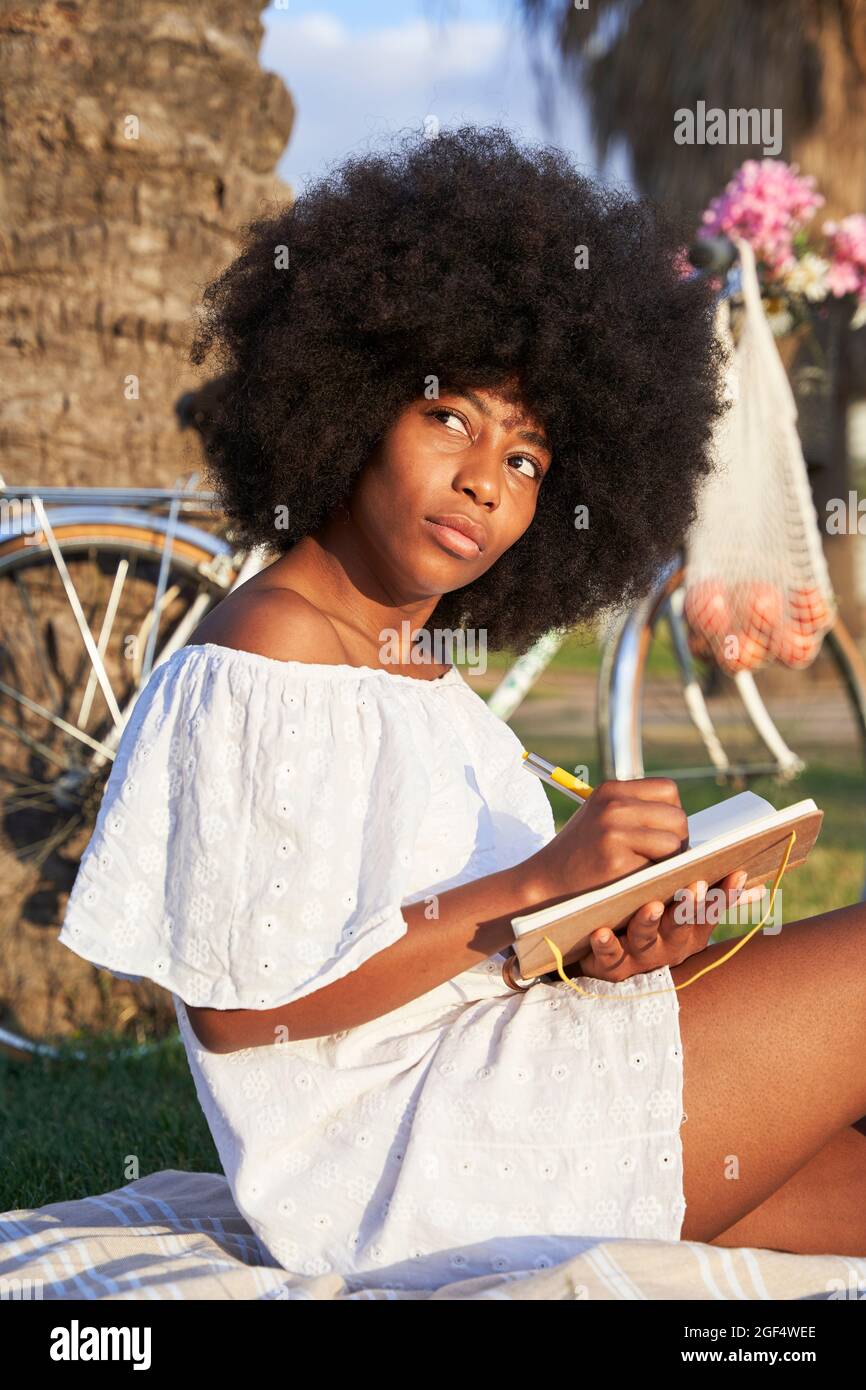 Afro woman contemplating while writing in book at park Stock Photo - Alamy