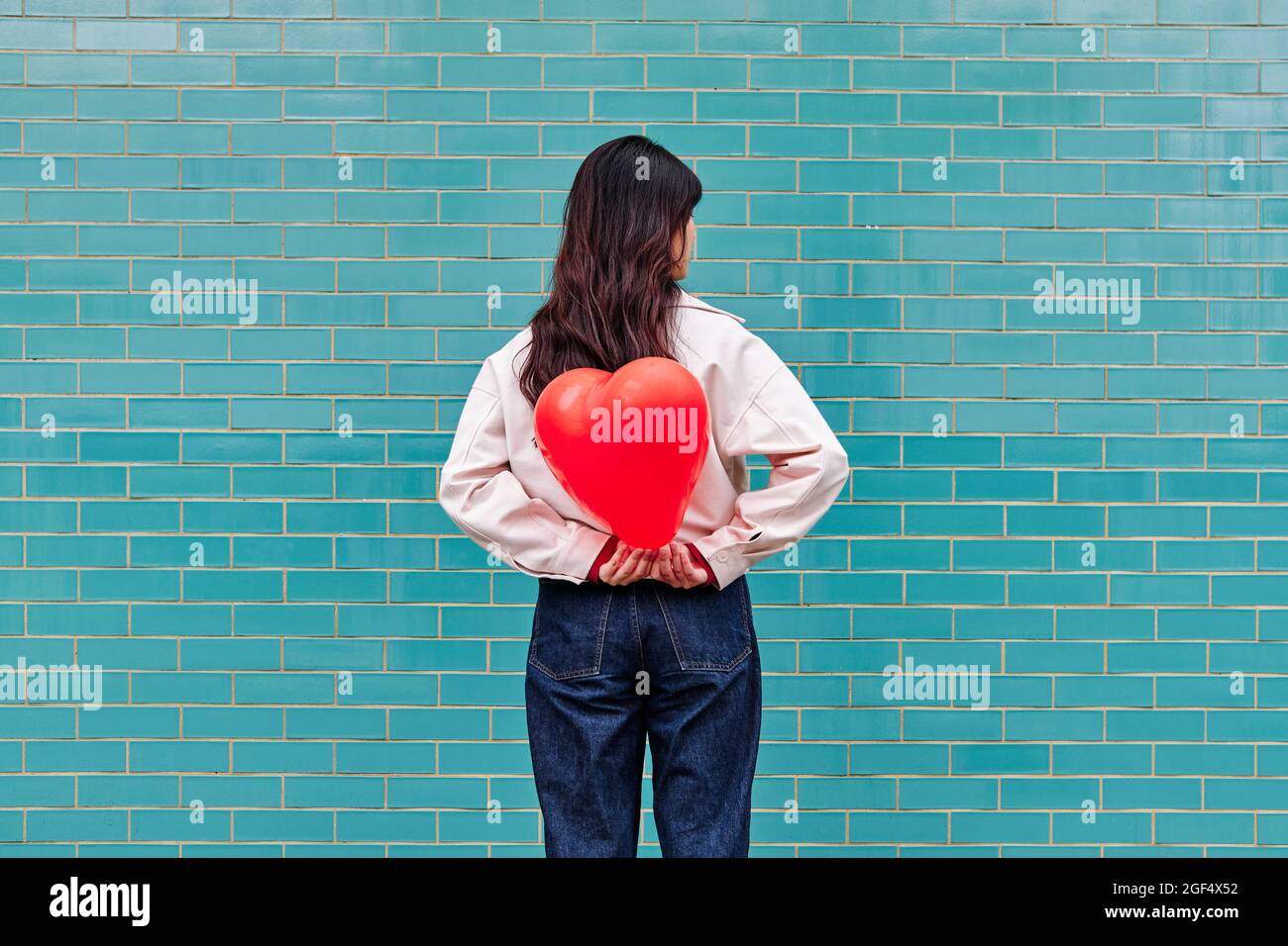 Woman holding heart shape balloon behind her back in front of turquoise ...