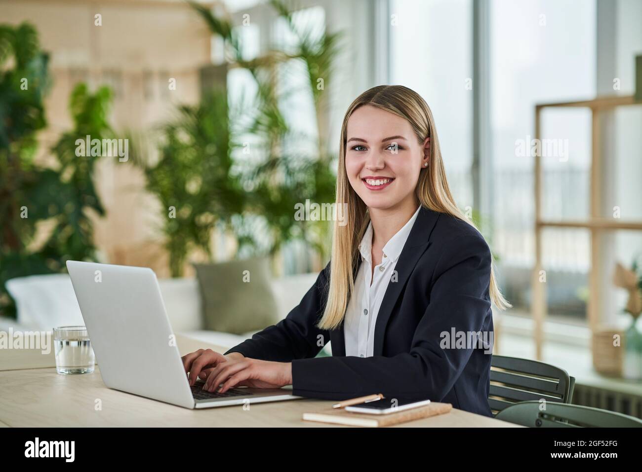 Smiling blond female trainee sitting with laptop at desk in office ...
