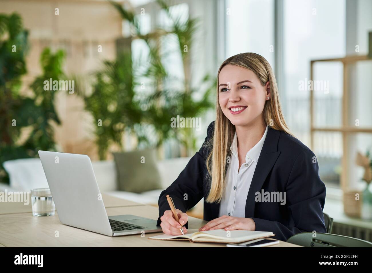 Happy blond teenage intern looking away while sitting with diary at ...