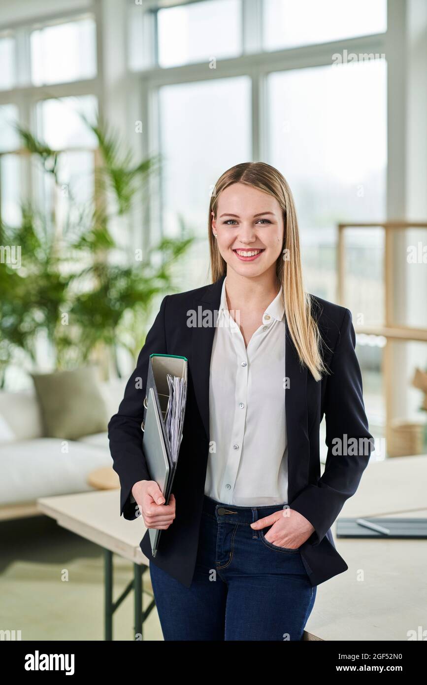 Smiling blond female intern holding file while standing with hand in ...