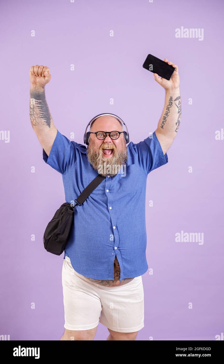 Excited fat man with headphones raises up hands and cellphone posing on ...