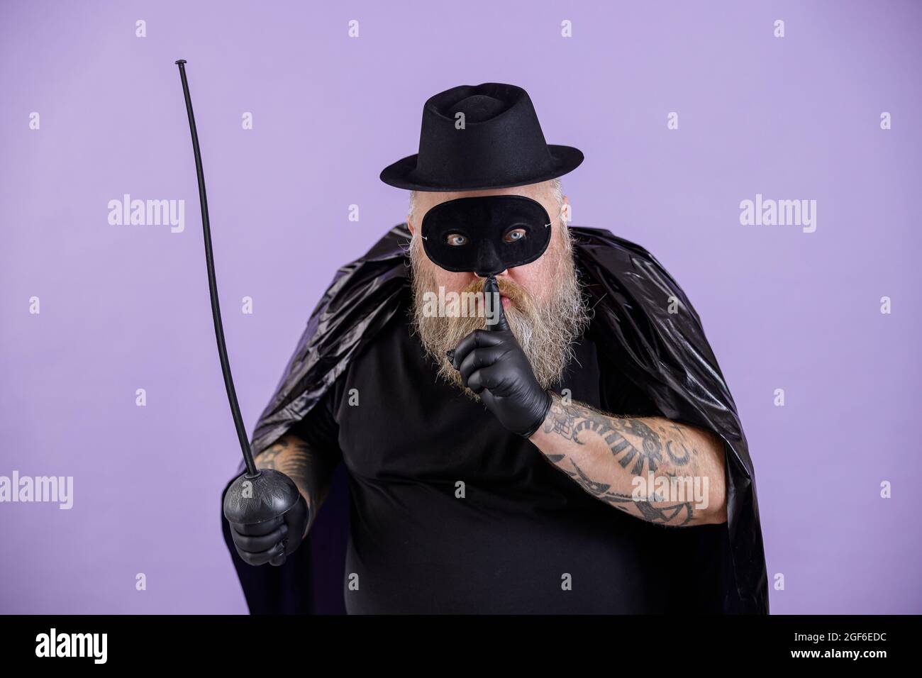 Bearded plump man in Zorro costume shows Hush gesture on purple ...