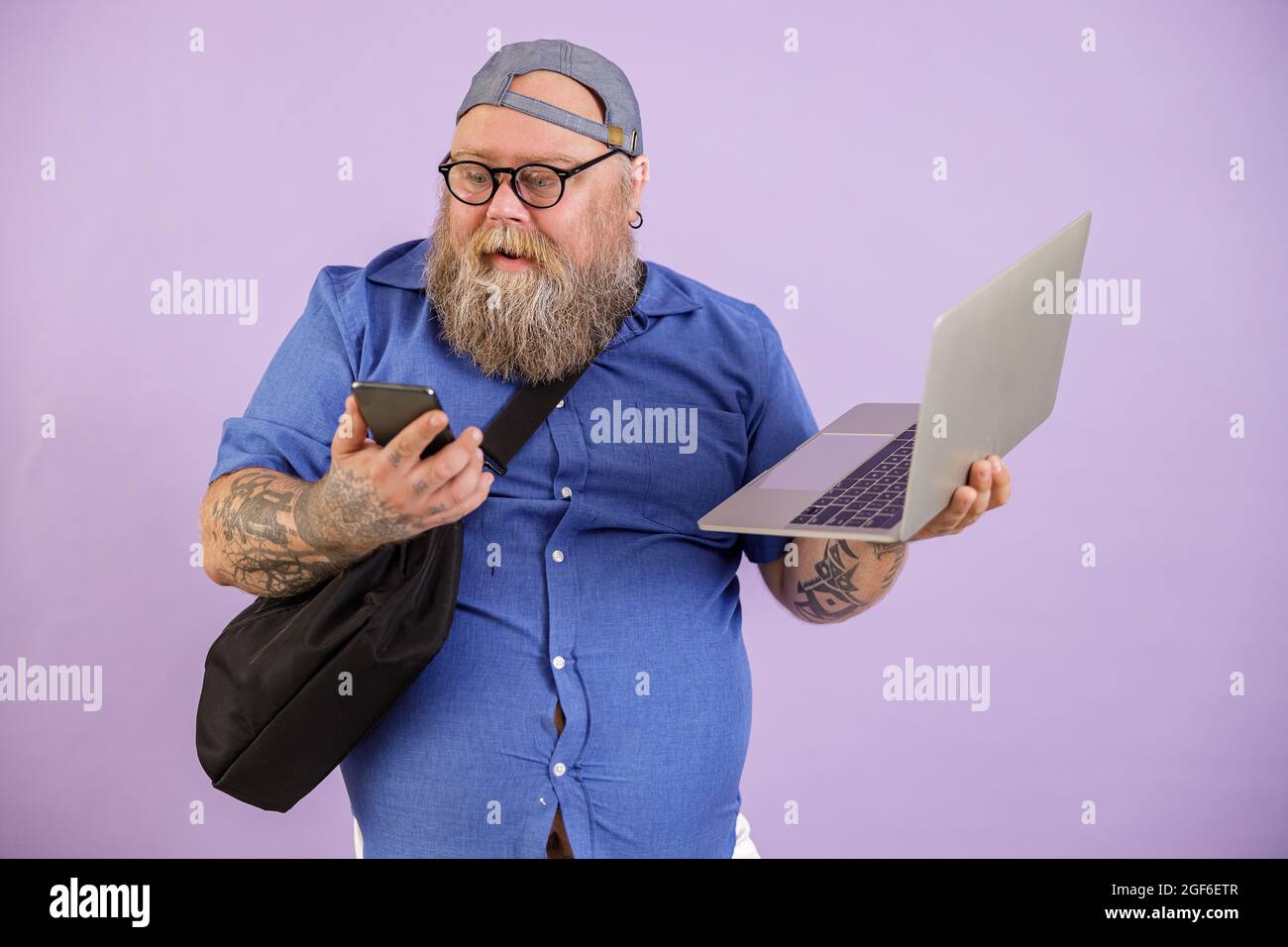 Surprised fat male person with laptop looks at mobile phone screen on ...