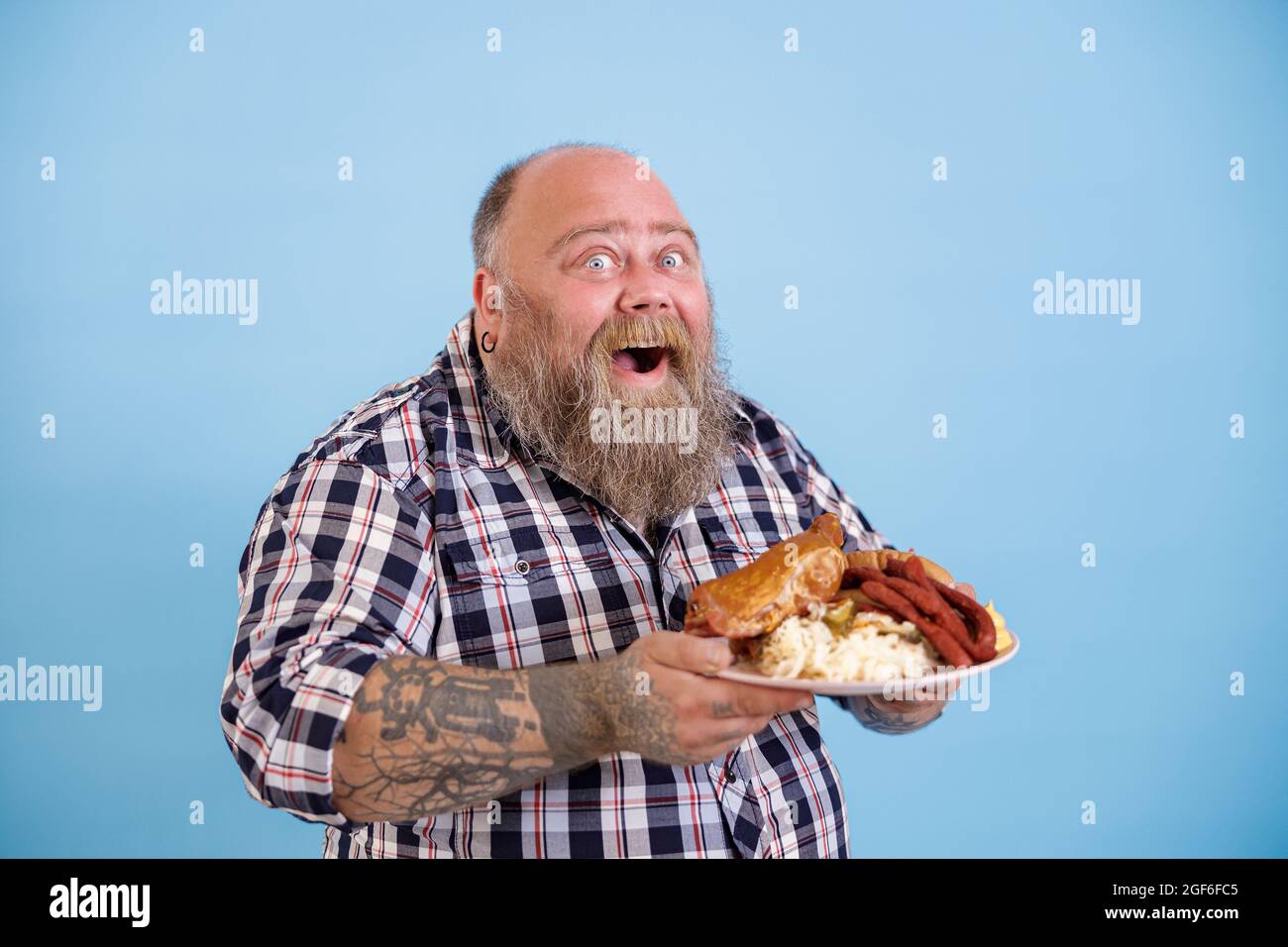 Happy plump man holds plate of fat food posing on light blue background ...