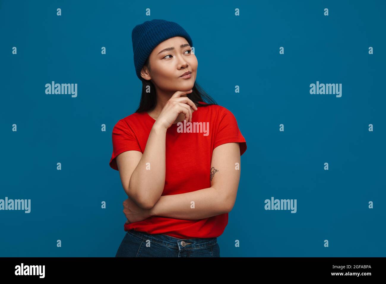 Asian teen girl in hat thinking and looking aside isolated over blue ...