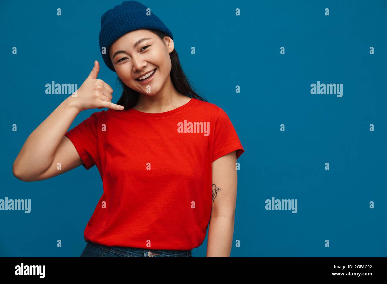 Asian teen girl in hat smiling and showing handset gesture isolated ...