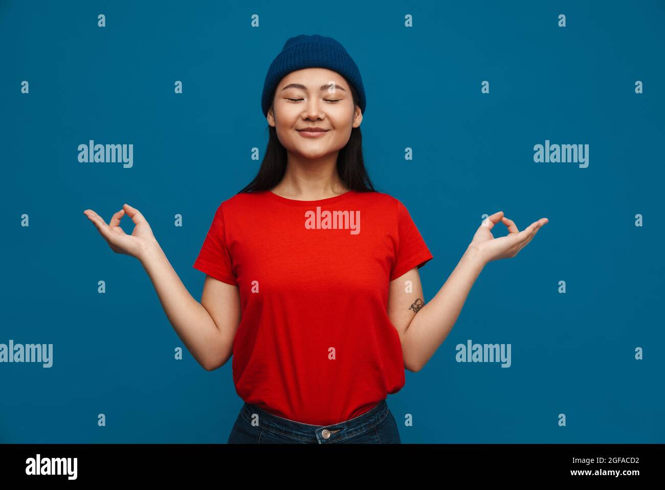 Asian teen girl in hat smiling and showing zen gesture isolated over ...