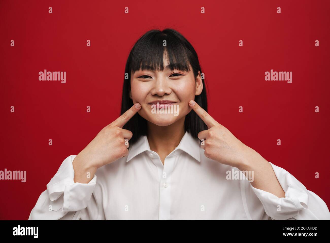 Young asian woman smiling and pointing fingers at her cheeks isolated ...
