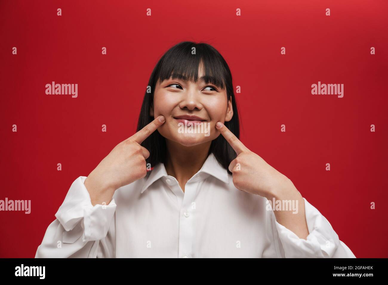 Young asian woman smiling and pointing fingers at her cheeks isolated ...