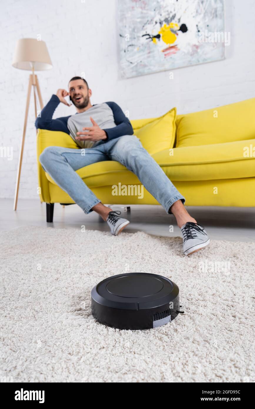 Robotic vacuum cleaner near blurred man talking on smartphone in living ...