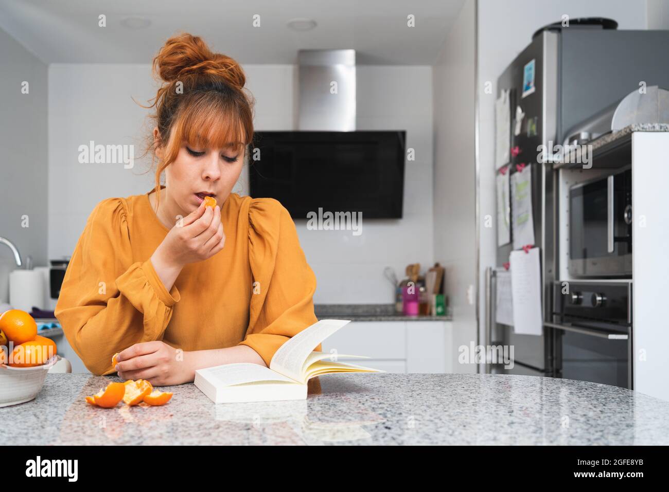 Caucasian female eating a tangerine while reading a book on a table ...