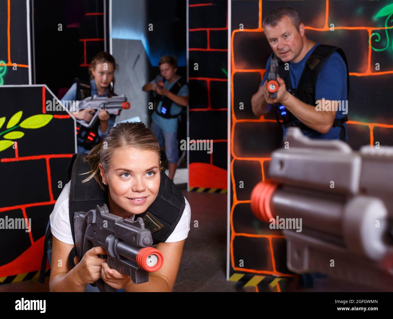 Smiling female aiming laser gun at other players during laser tag game ...