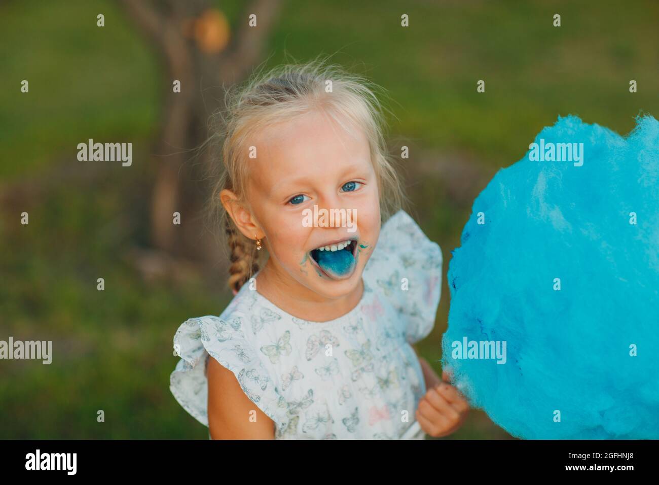 Little blond girl eating cotton candy and shows blue tongue in the park ...