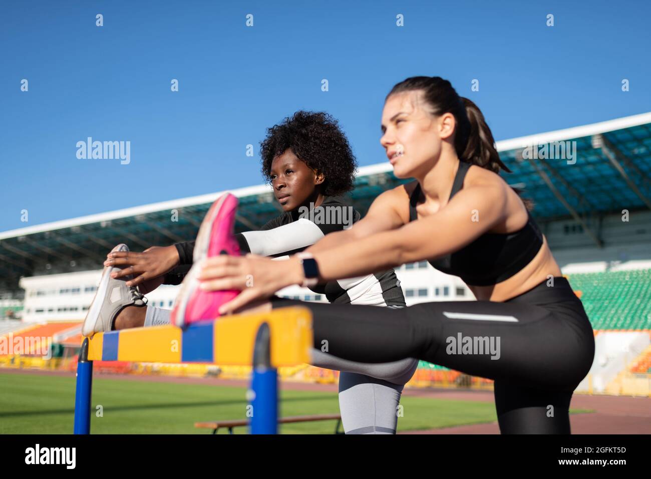 Focused diverse female athletes stretching with legs on barrier while ...
