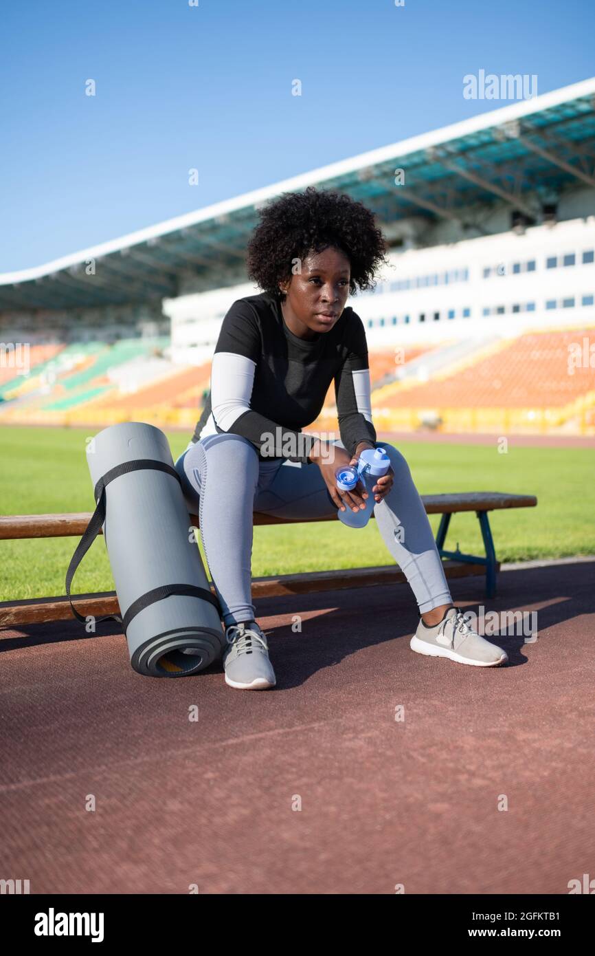Black female athlete with water sitting on bench during break on ...