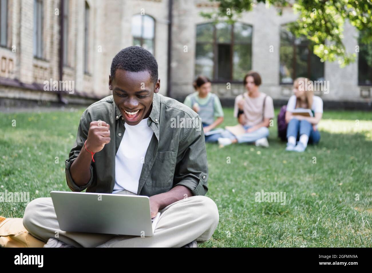 Excited african american student showing yes while using laptop on lawn ...