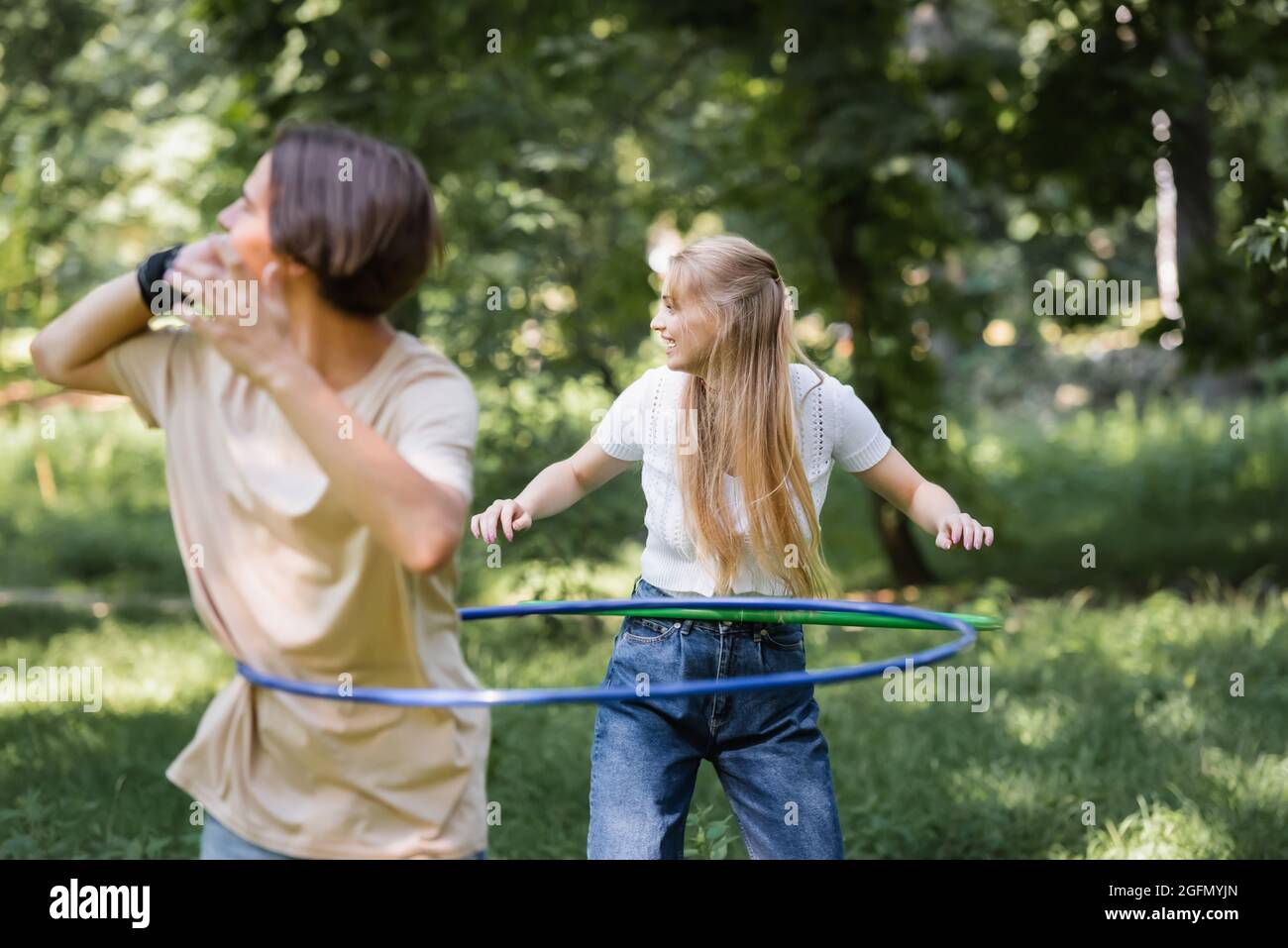 Side view of smiling girl twisting hula hoop near blurred friend in ...