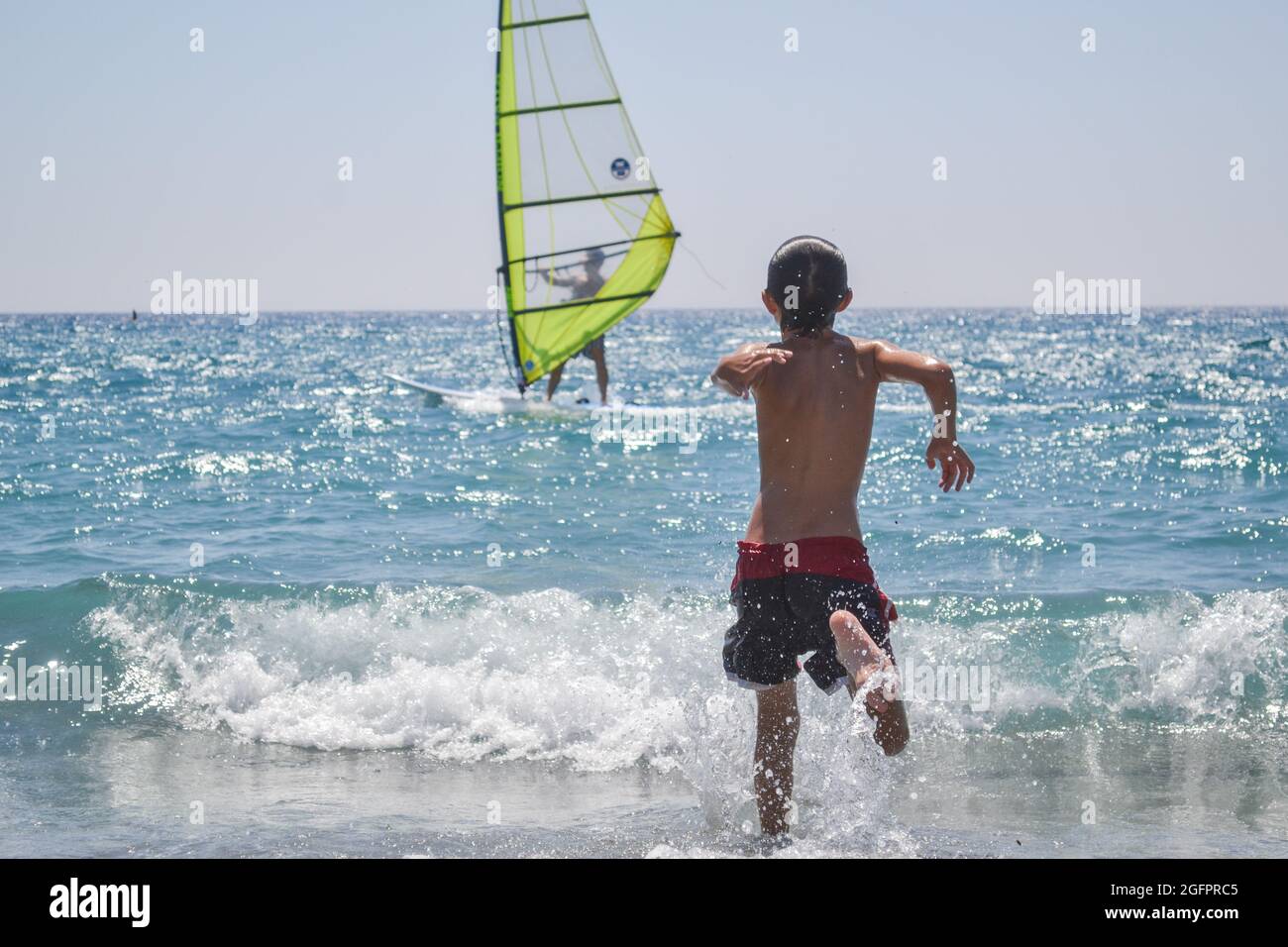 Boy running into the sea and in the background a surfboard with sail ...
