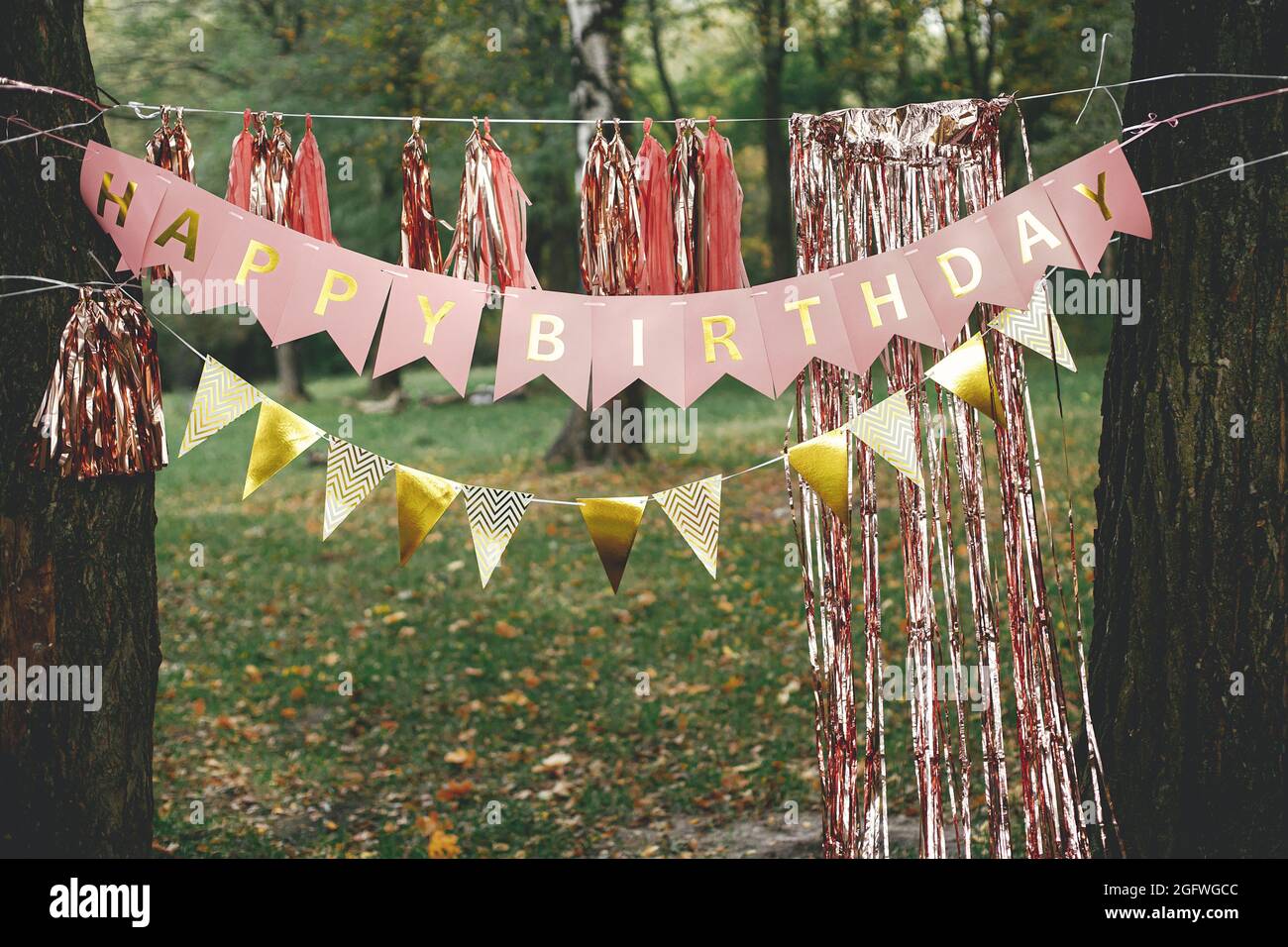 Stylish happy birthday garland hanging in park. Modern rose gold decor ...