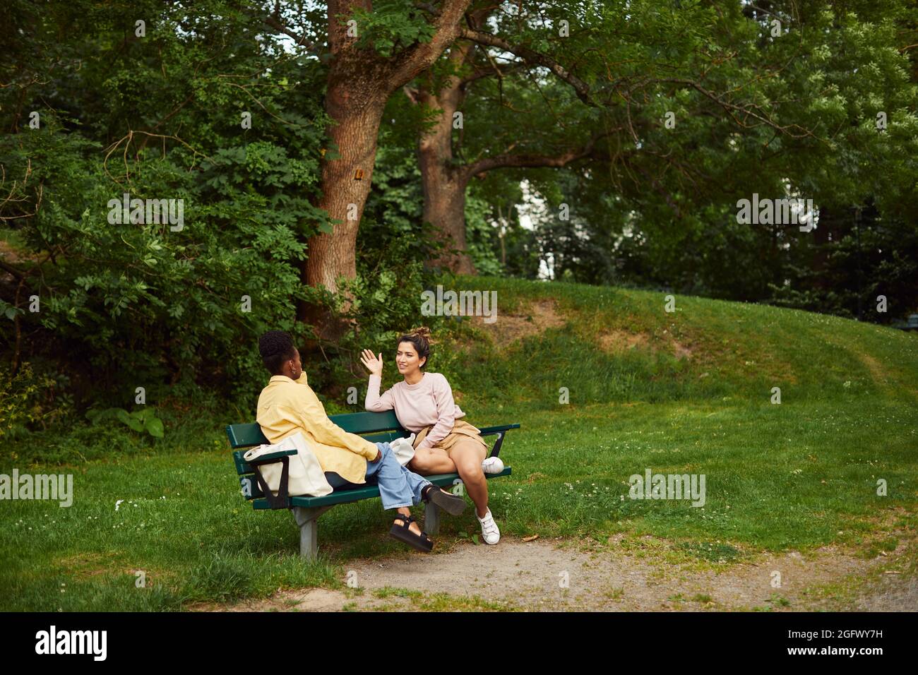 Female friends sitting on bench and talking Stock Photo - Alamy