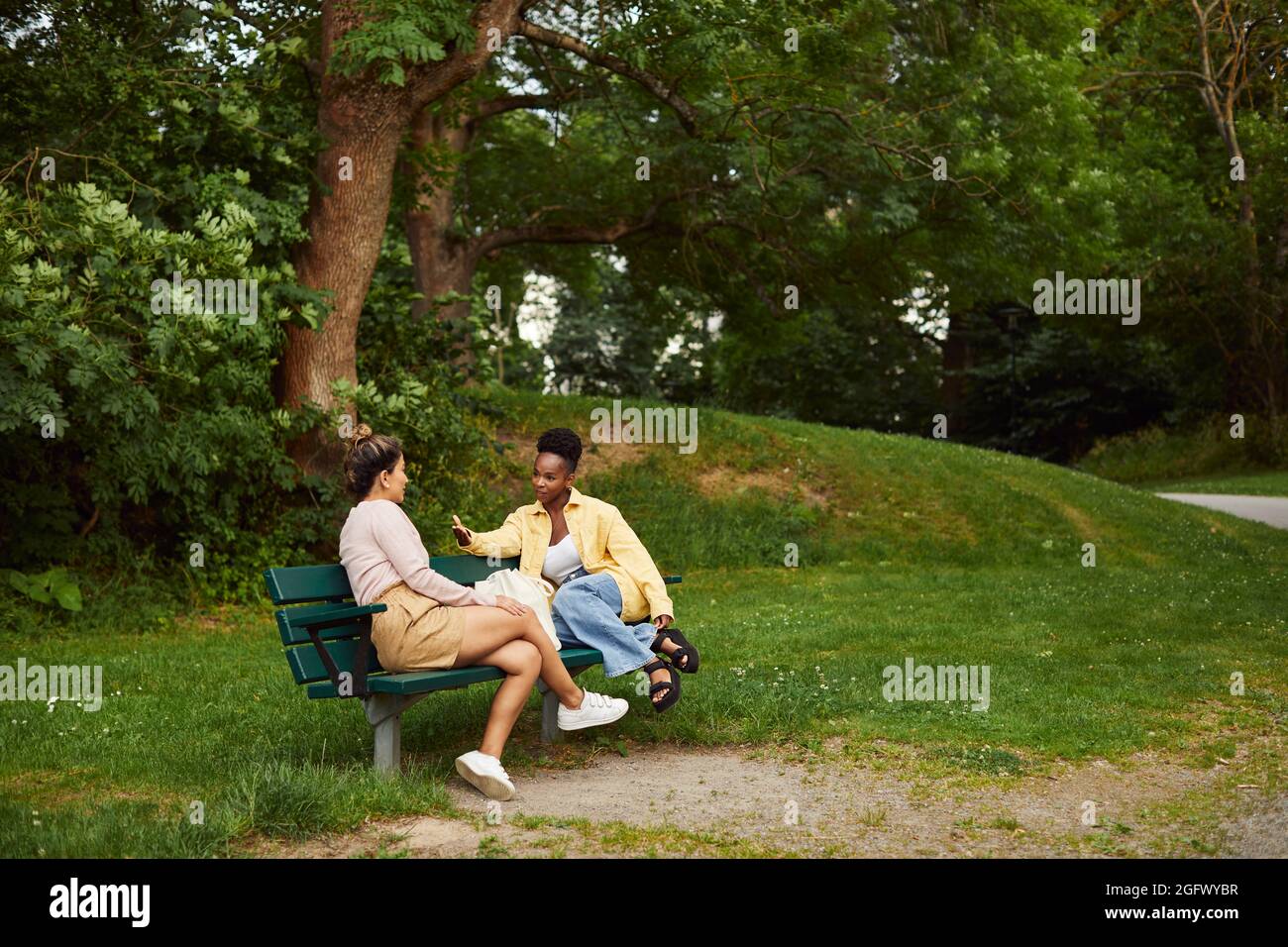 Female friends sitting on bench and talking Stock Photo - Alamy