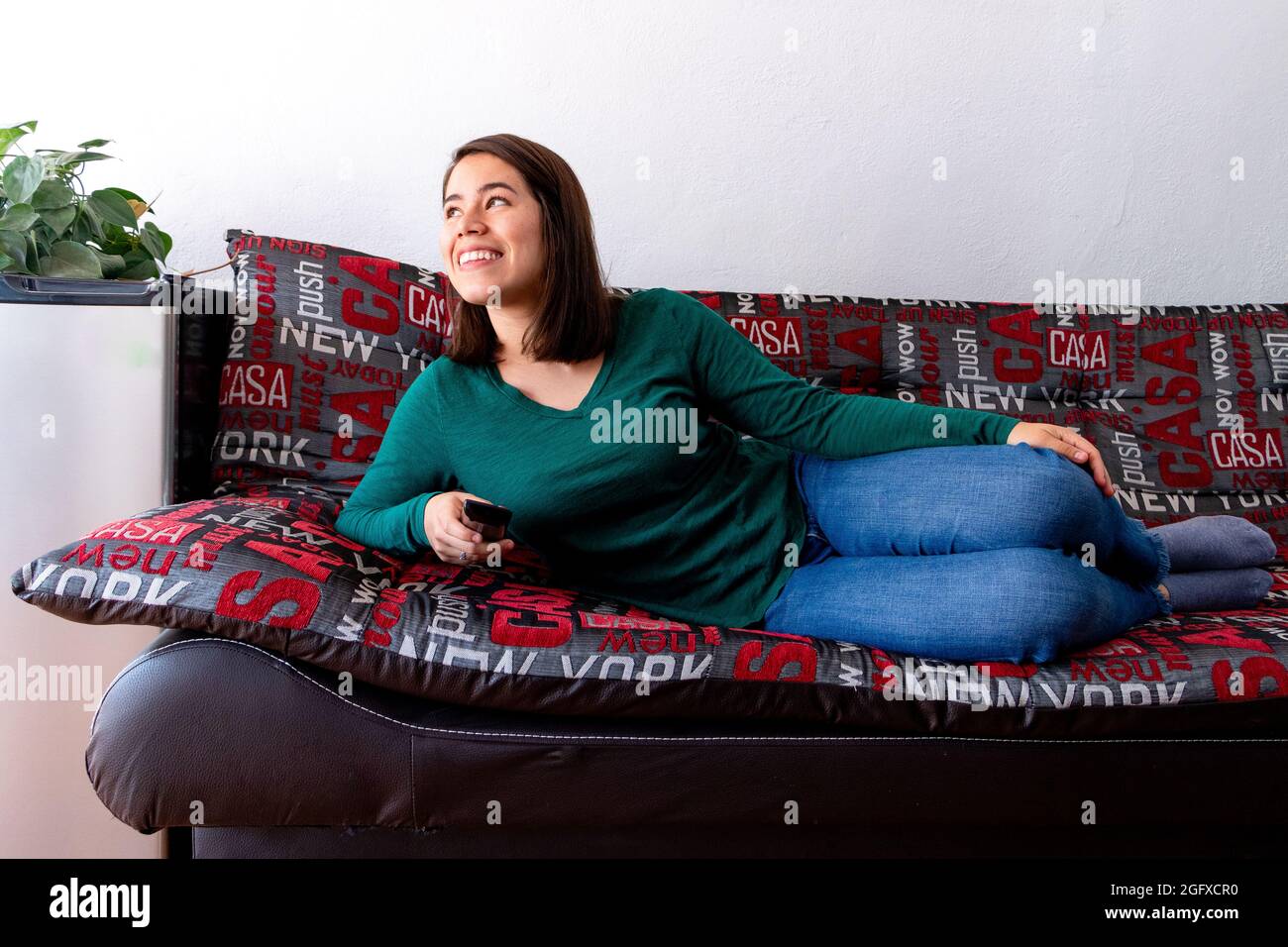 Cheerful young Hispanic lady lying on the couch holding a remote ...