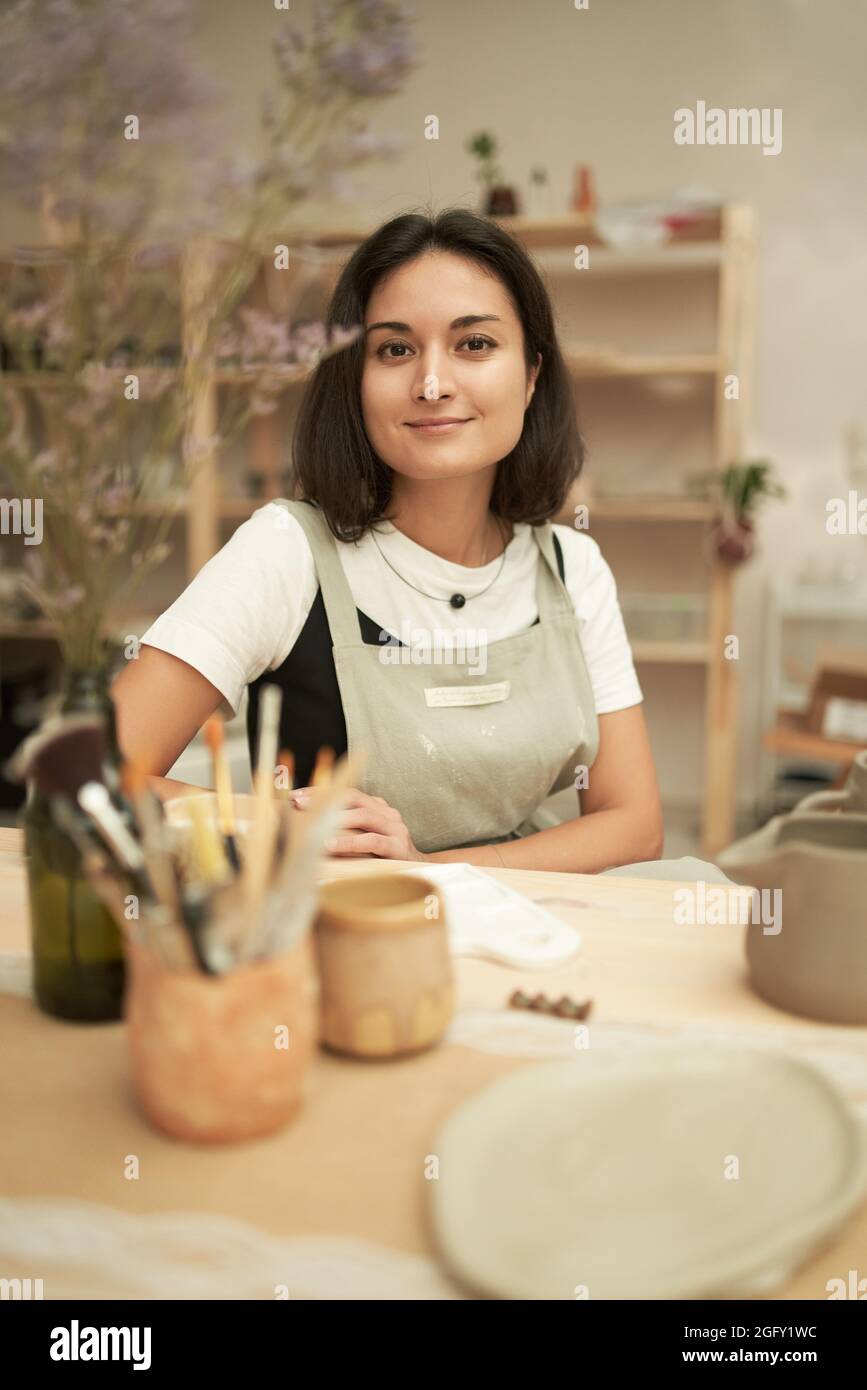 Young craftswoman in apron sitting at table and looking at camera ...