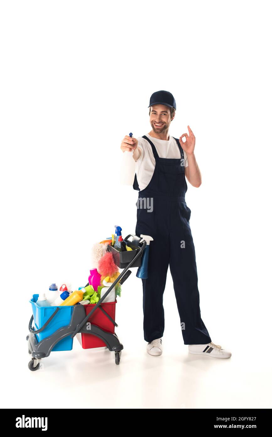 Smiling cleaner holding detergent and showing ok near cart on white ...