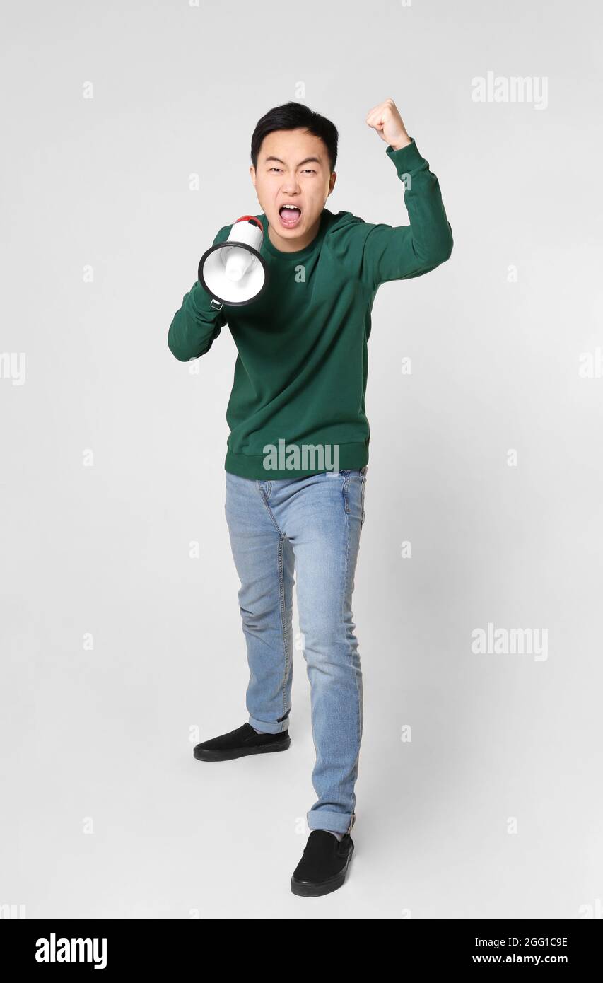 Protesting young Asian man shouting into megaphone on light background ...