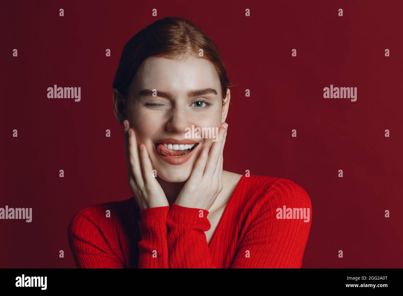 Portrait of young ginger funny woman winks on red background with hands ...