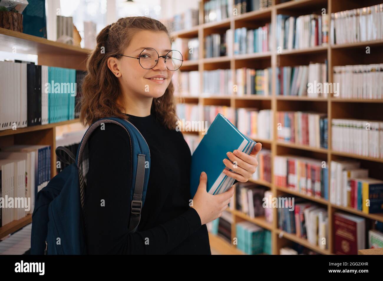 Happy teenage girl or student wearing glasses taking book from shelf in ...
