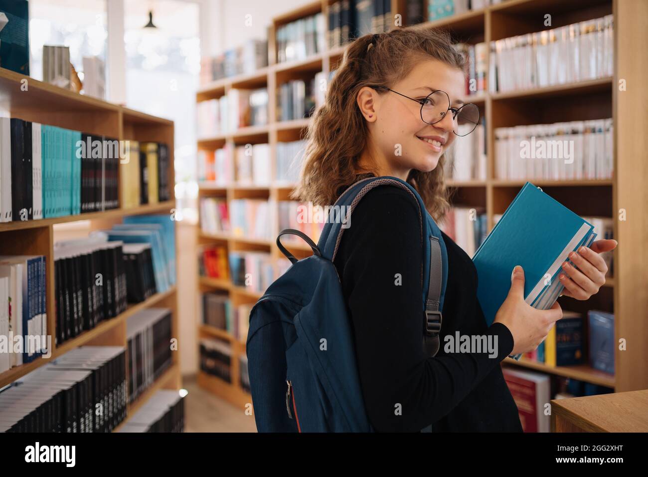 Happy teenage girl or student wearing glasses taking book from shelf in ...