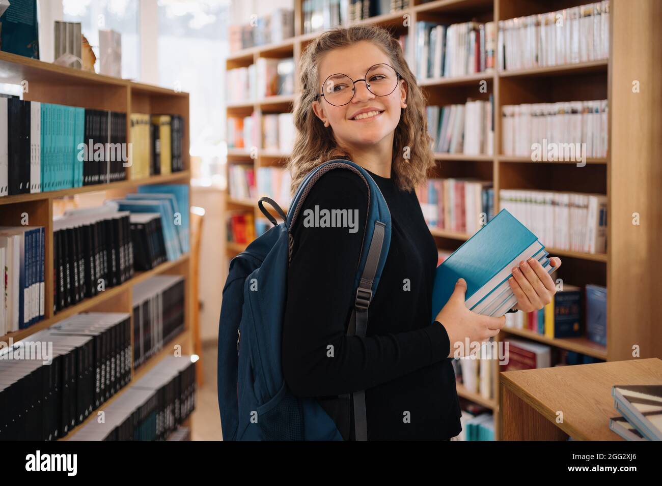Happy teenage girl or student wearing glasses taking book from shelf in ...