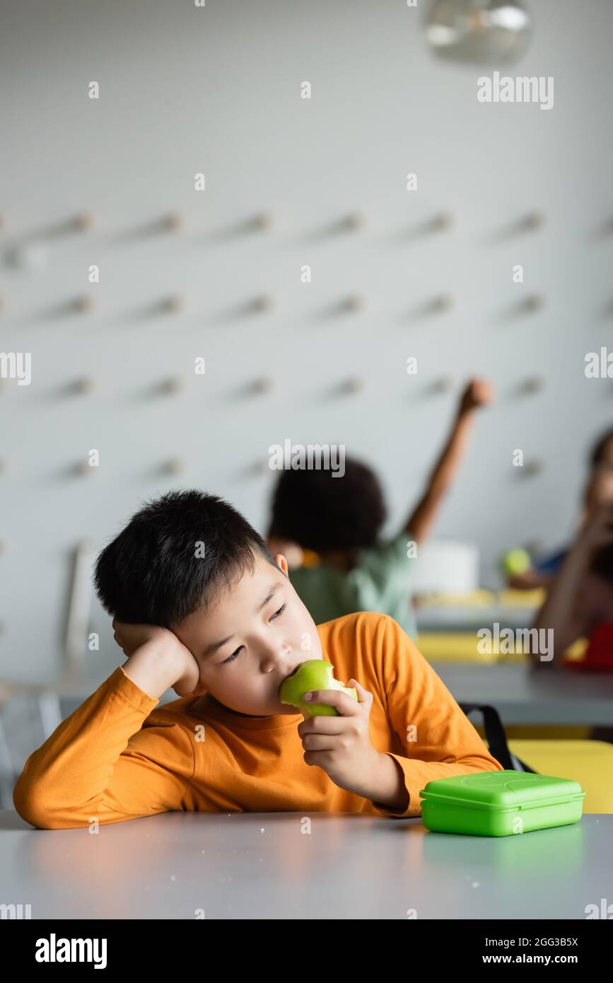 sad asian boy eating apple in school eatery near kids on blurred ...
