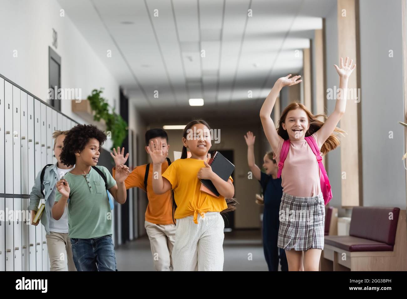 happy multiethnic pupils waving hands while running in school corridor ...