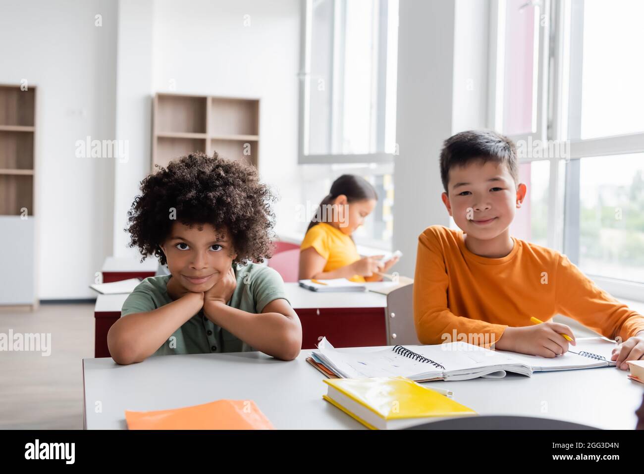 multiethnic classmates looking at camera near notebooks in classroom ...