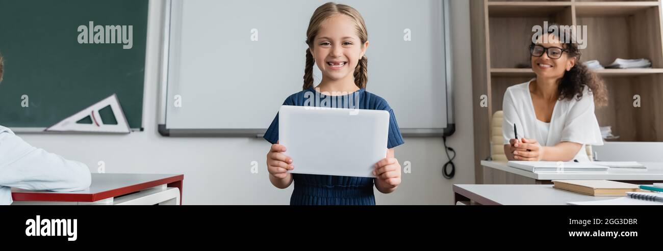 happy schoolgirl holding digital tablet near teacher smiling in ...