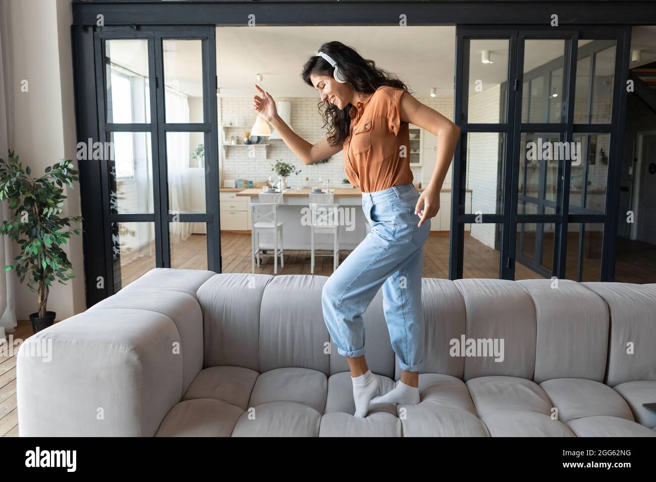 Positive young woman with headset dancing on couch while listening to ...