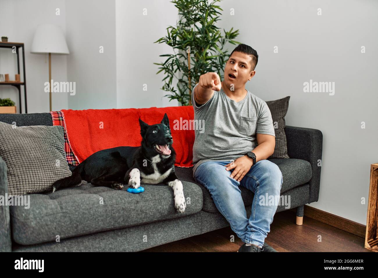 Young latin man and dog sitting on the sofa at home pointing displeased ...