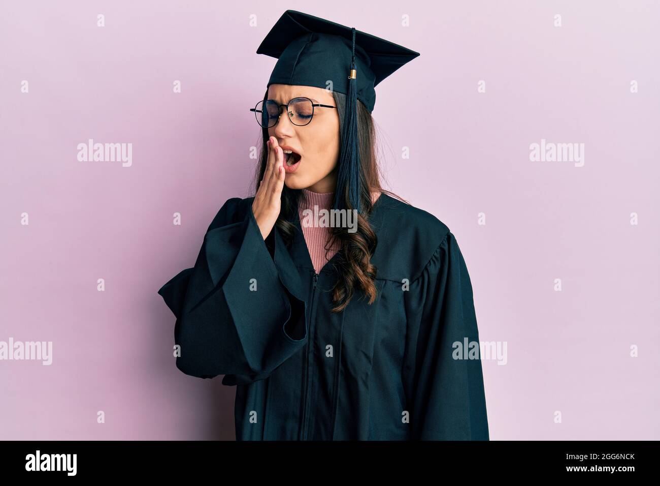 Young hispanic woman wearing graduation cap and ceremony robe bored ...