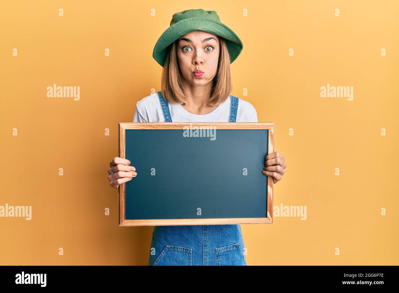 Young caucasian blonde woman holding blackboard puffing cheeks with funny face. mouth inflated ...