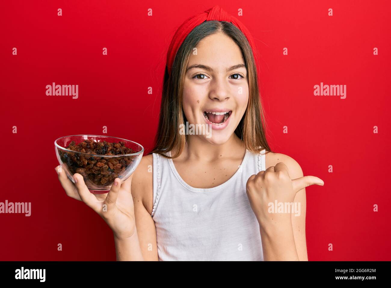 Beautiful brunette little girl holding bowl of raisins pointing thumb ...